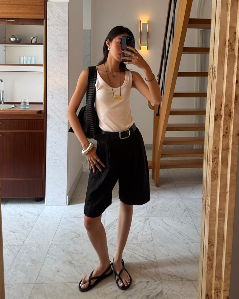 White tank top with tailored black Bermuda shorts%2C leather sandals%2C woven shoulder bag and gold jewelry styled in modern interior mirror