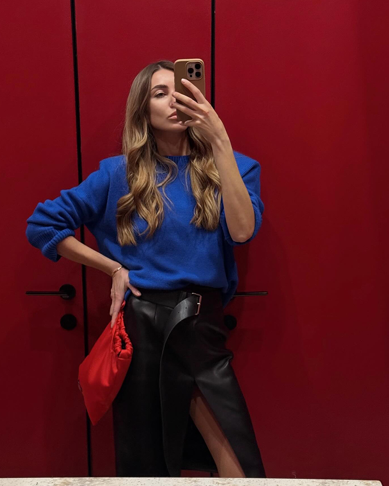 Woman in blue knit sweater and black leather wrap skirt with red clutch%2C styled in bold red interior mirror selfie setting
