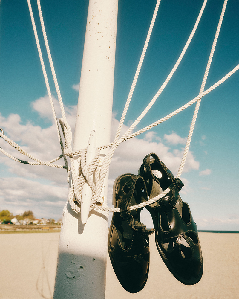 Black leather fisherman sandals hanging from sailboat ropes against a sunny beach backdrop for a nautical summer look