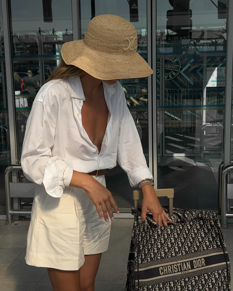Chic summer look with white button-down shirt and high-waisted shorts%2C styled with straw hat and Christian Dior tote at airport entrance