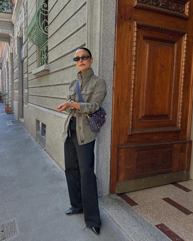 Woman in olive military jacket and black wide-leg trousers with monogram shoulder bag%2C styled on a European city street by stone facade