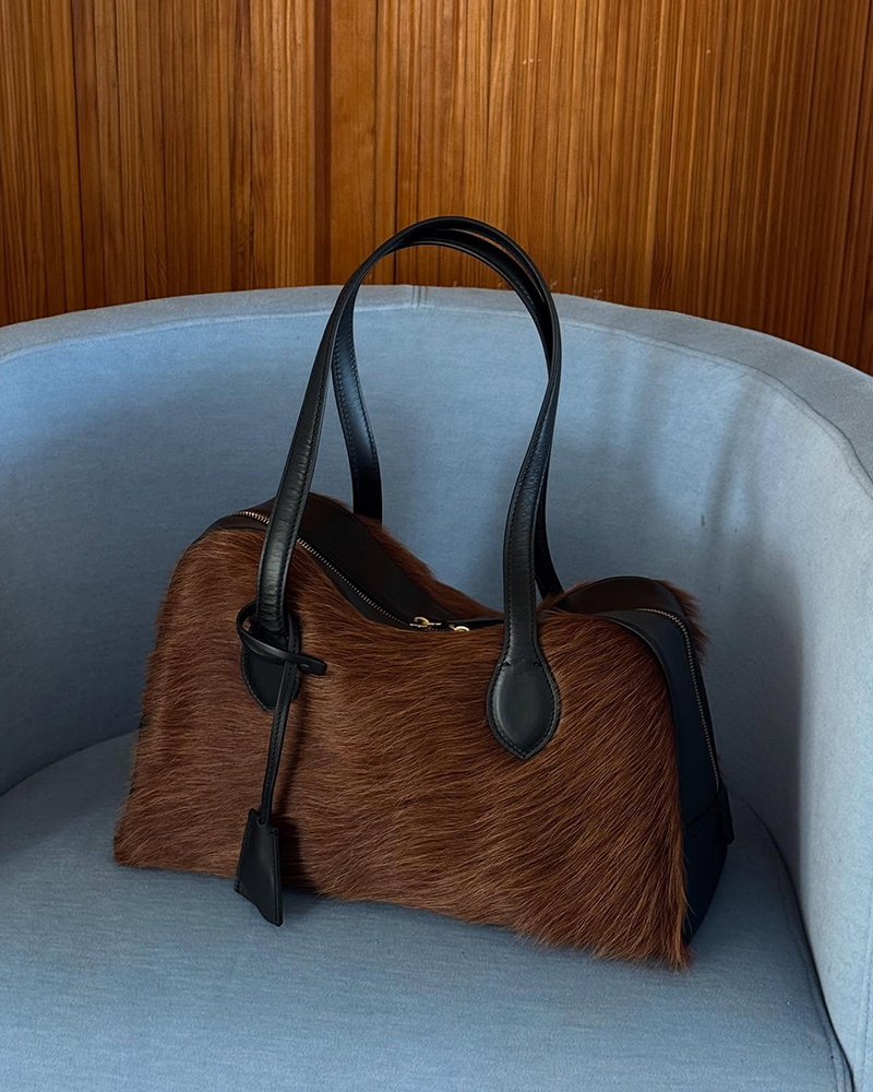 Brown textured calf hair top-handle handbag with black leather straps and gold hardware%2C styled on a pale blue armchair indoors