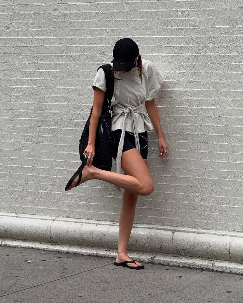 Wrap-front linen top paired with black biker shorts%2C flip-flops%2C baseball cap and quilted tote against a minimalist city wall