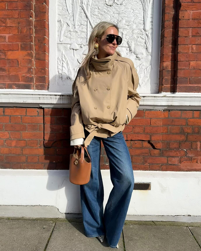 Woman in beige cropped trench jacket and wide-leg blue jeans with leather bucket bag and sunglasses%2C posed against brick wall