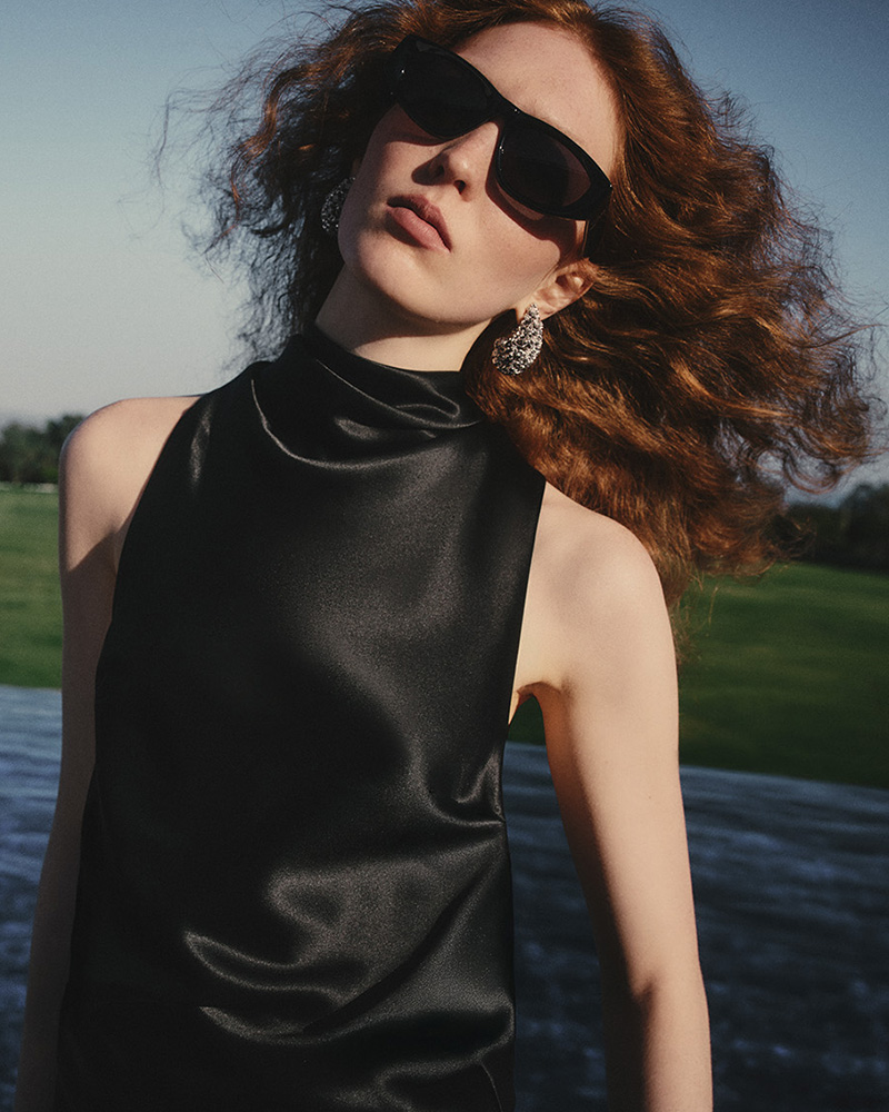 Woman in black satin halter top with oversized sunglasses and statement earrings%2C posed outdoors against a sunlit landscape