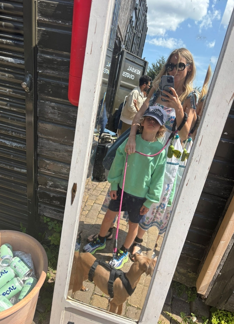 Mirror selfie of mother in printed sundress with tote and sneakers walking child and dog outdoors