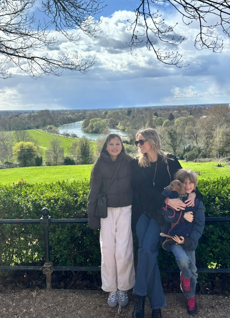 Mother in black coat with jeans and boots holding children and dog, set against a scenic countryside view
