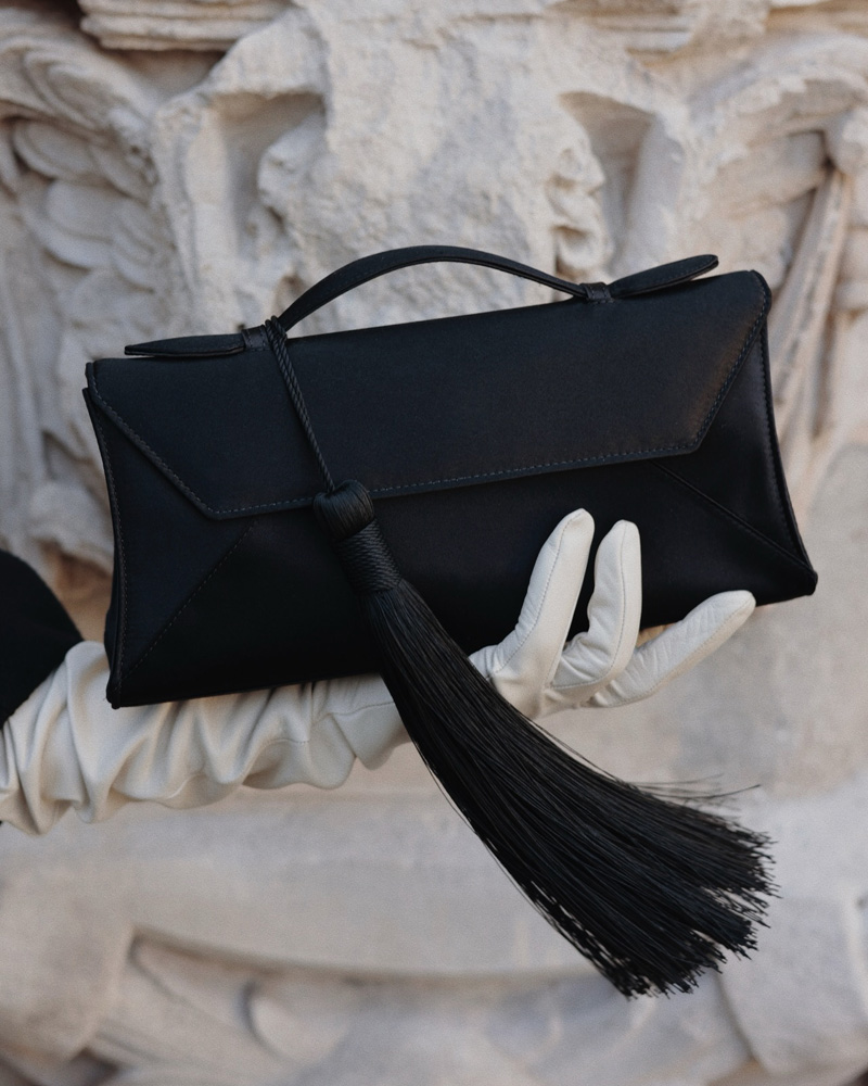 Black leather clutch bag with tassel detail and top handle%2C styled with white gloves against a textured stone backdrop