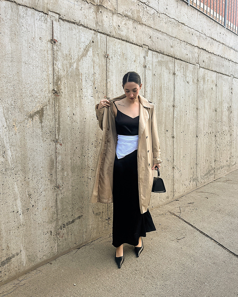 Woman in black slip dress with white scarf belt and beige trench coat%2C carrying mini bag on minimalist city sidewalk