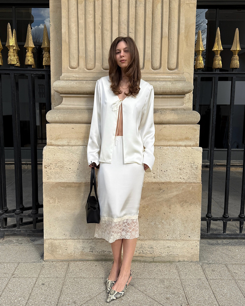 Cream silk blouse with lace-trim midi skirt%2C slingback heels and mini handbag styled against classic stone building