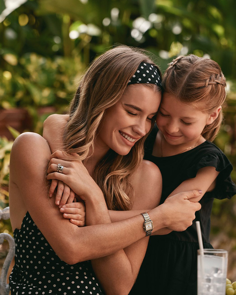 Mother and daughter in black polka dot dress and black dress with headband and watch%2C sharing a moment in lush garden setting