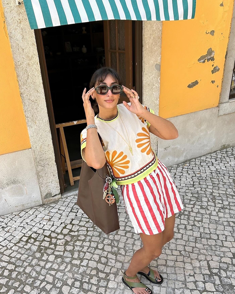 Woman in floral print tee and red striped shorts with sunglasses%2C green sandals and brown tote on cobblestone street by yellow facade