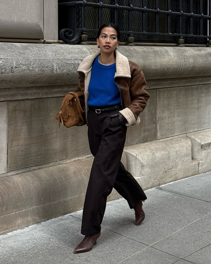 Model in a brown shearling jacket%2C blue sweater%2C dark trousers and leather boots with a suede bag on a city sidewalk