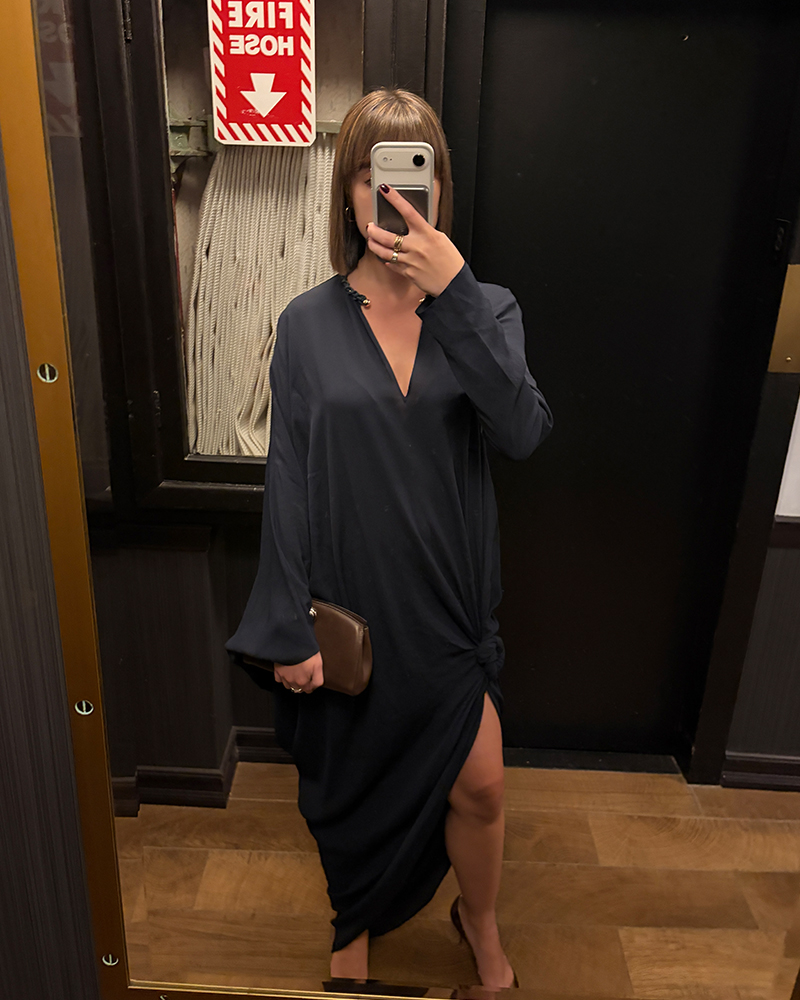 Woman wearing a navy blue long-sleeve midi dress with a side knot and slit%2C holding a brown leather clutch and wearing gold rings