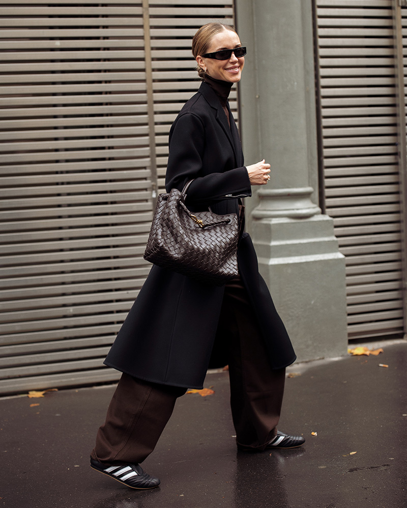 Street style look with black tailored coat%2C brown wide-leg trousers%2C woven leather shoulder bag and Adidas sneakers on city sidewalk