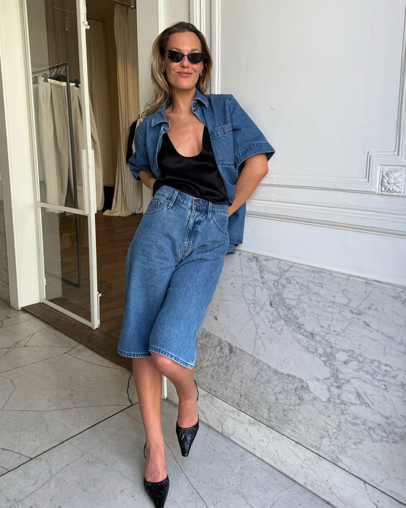 Woman in denim shirt and Bermuda shorts with black satin tank%2C pointed heels and sunglasses%2C styled in minimal indoor setting