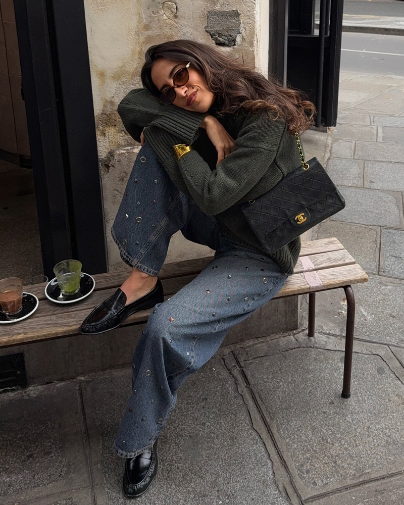 Woman in embellished blue jeans with green knit sweater%2C Chanel quilted shoulder bag%2C loafers%2C and sunglasses%2C seated at caf%C3%A9 bench