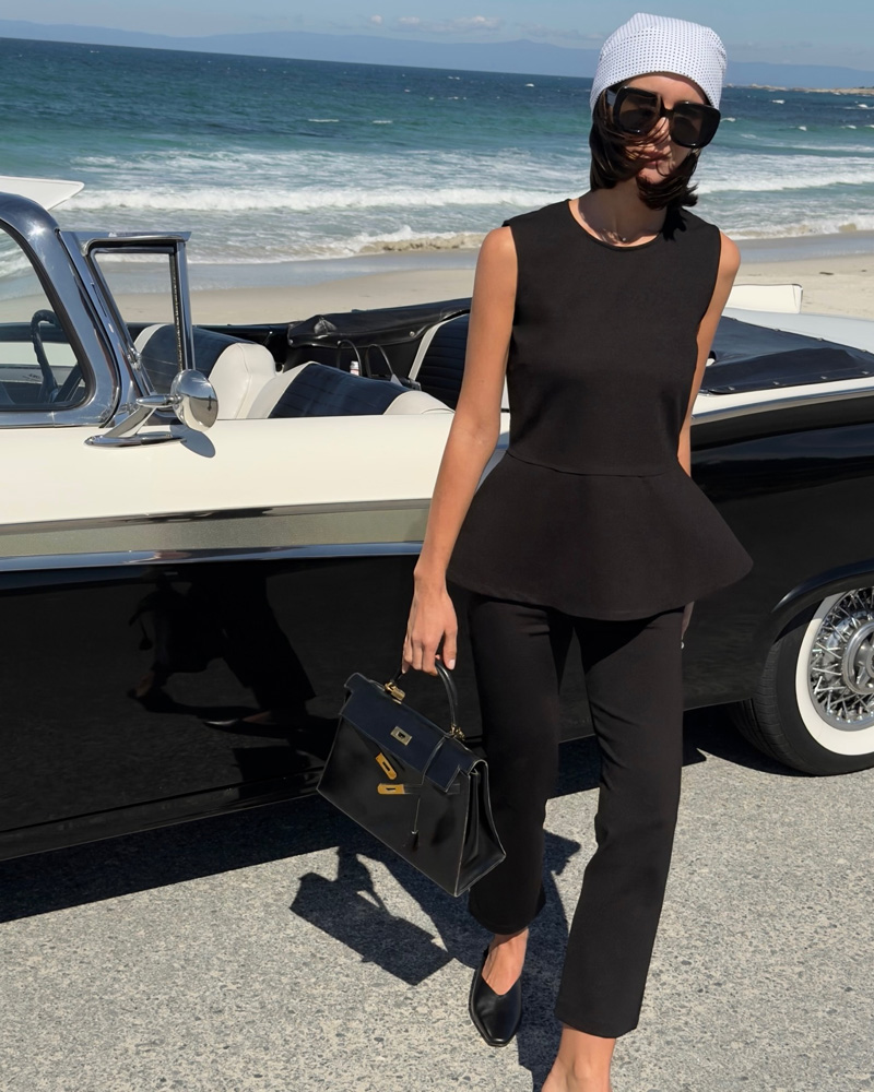 Woman in black peplum top and slim pants with structured leather handbag and sunglasses%2C styled by vintage car on beach road
