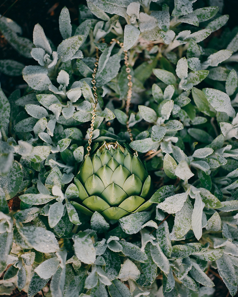 Green artichoke-shaped novelty handbag with gold chain strap nestled in soft sage foliage outdoors