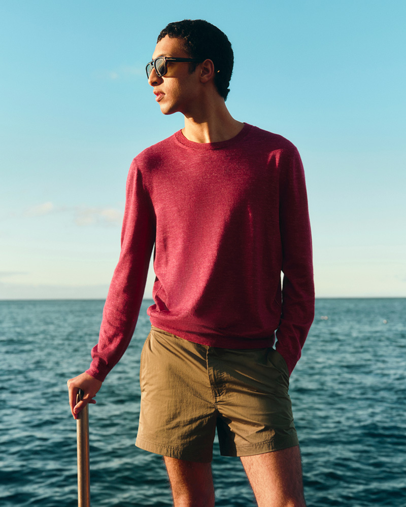Man in red lightweight knit sweater with khaki shorts and sunglasses%2C standing by ocean in breezy summer seaside setting