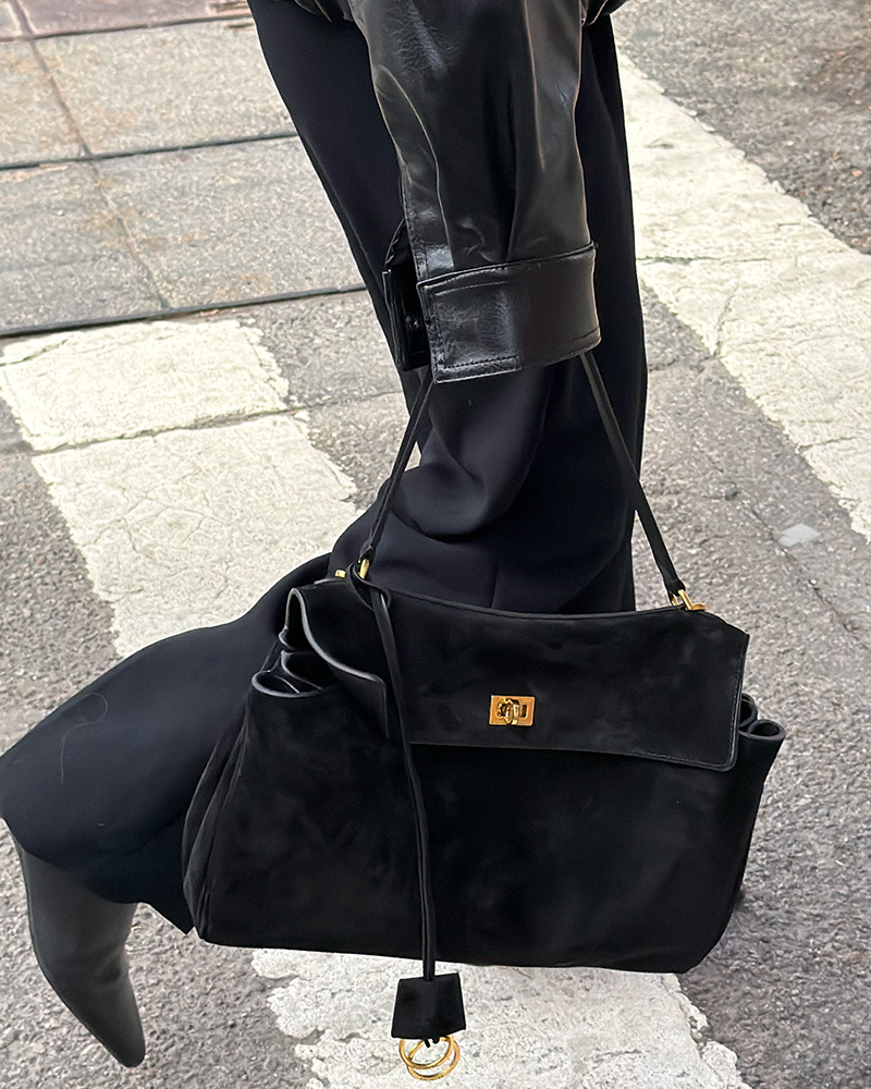 Close-up of black leather shoulder bag with gold hardware styled with sleek black boots on a city crosswalk