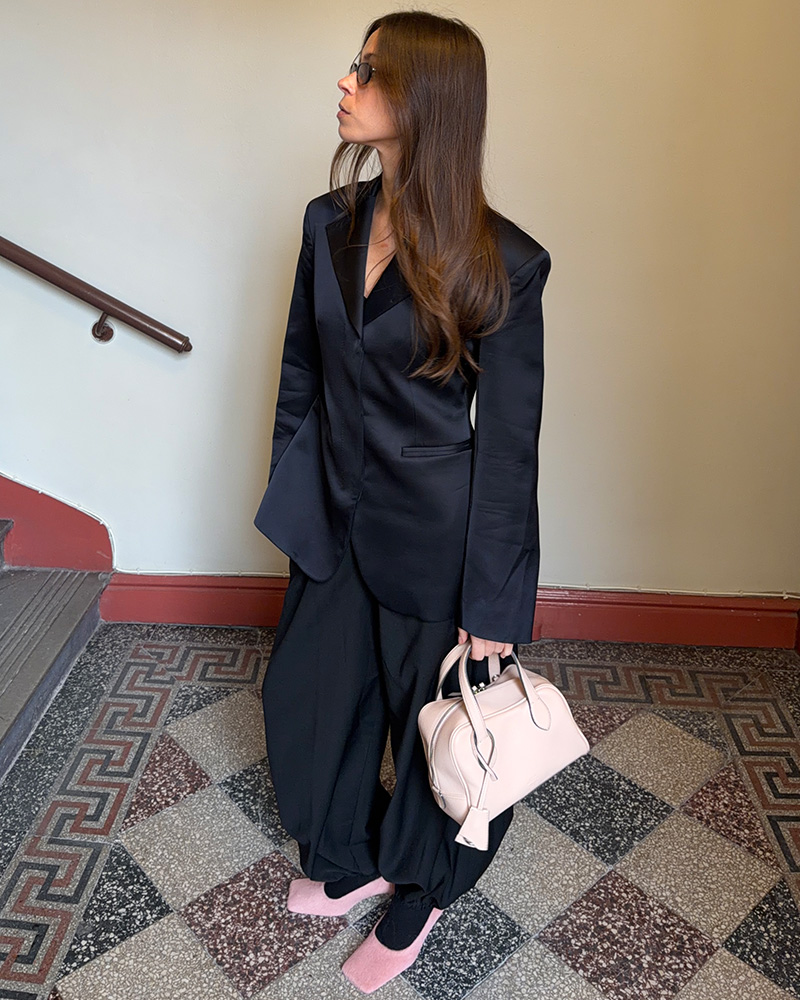 Model wearing a black satin blazer with wide-leg trousers%2C pink square-toe shoes%2C and a light pink leather handbag in a minimalist indoor setting