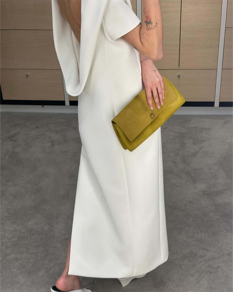 Woman in minimalist white draped dress holding a mustard suede clutch%2C styled with black heels in a modern indoor setting