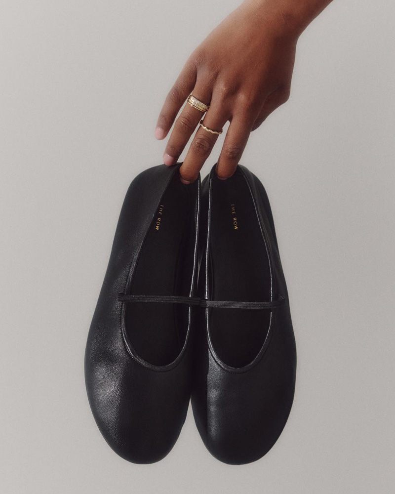The Row black leather Mary Jane flats with elastic strap%2C held by hand with gold rings against minimal neutral backdrop
