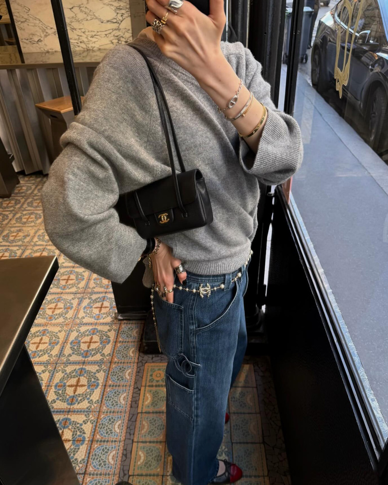 Chanel mini flap shoulder bag styled with gray knit sweater%2C blue jeans%2C and layered gold jewelry in chic caf%C3%A9 setting