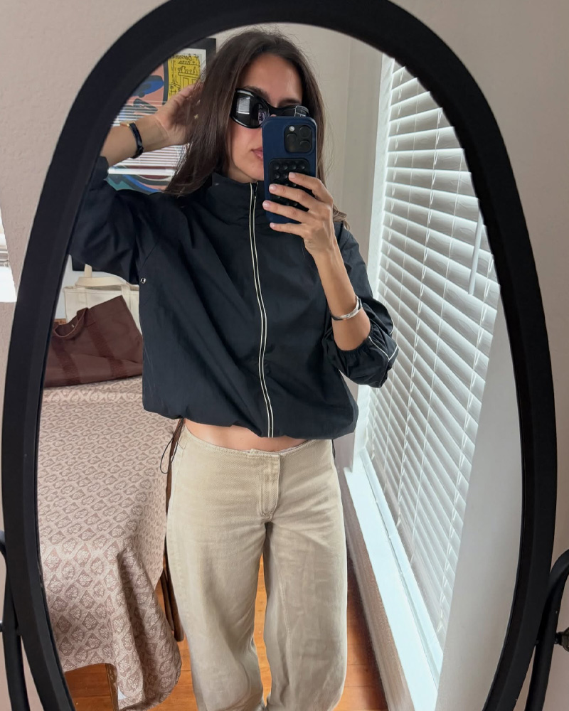 Woman in black cropped technical jacket with beige trousers and sunglasses%2C taking mirror selfie in sunlit minimalist interior
