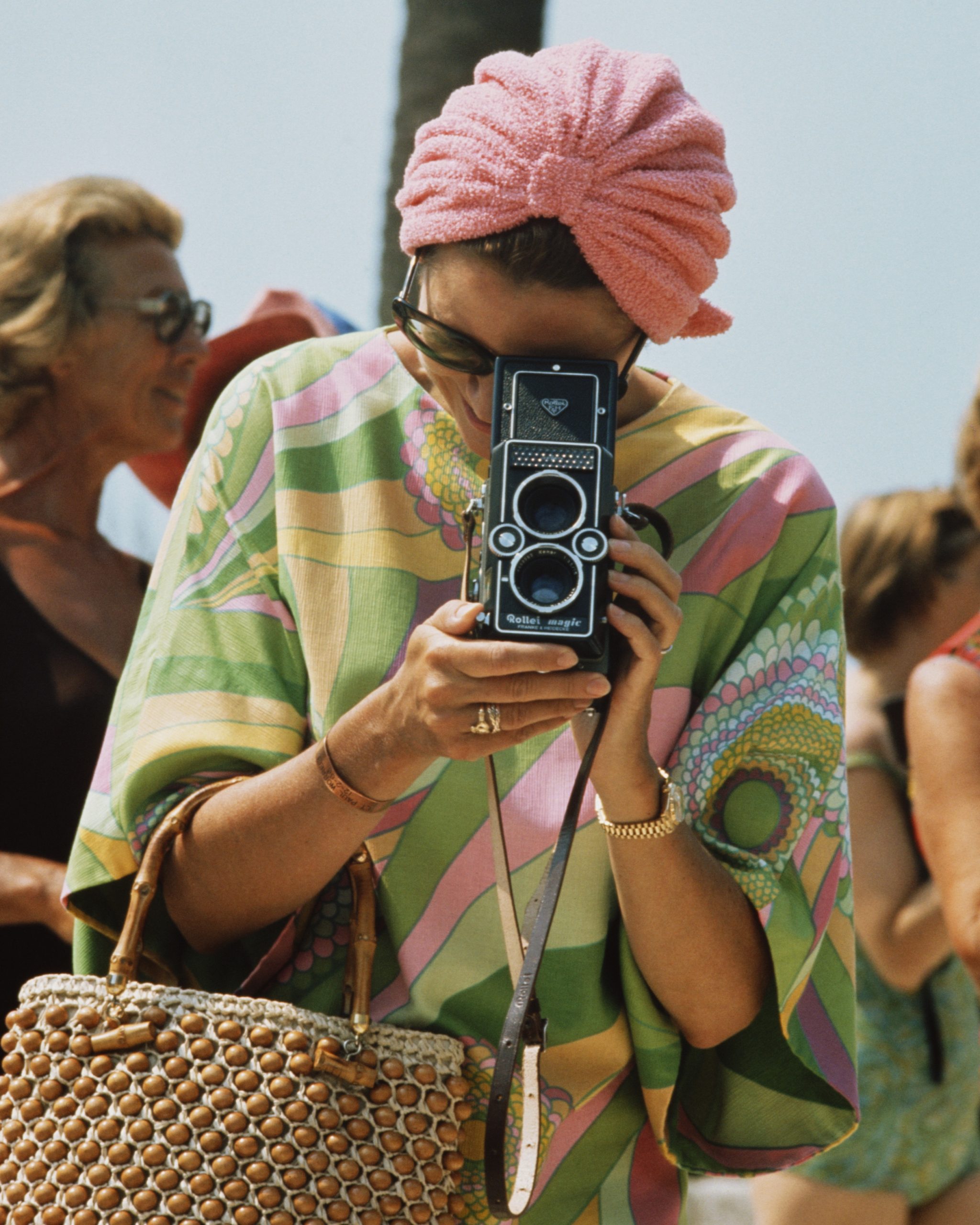 Woman wearing a colorful vintage-inspired kaftan with green%2C pink%2C and yellow patterns%2C a pink terry cloth turban%2C and holding a retro Rolleiflex camera with a woven bamboo handbag