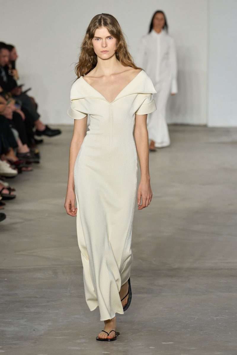 Model walking runway in a cream off-shoulder maxi dress with short sleeves and black sandals at a fashion show