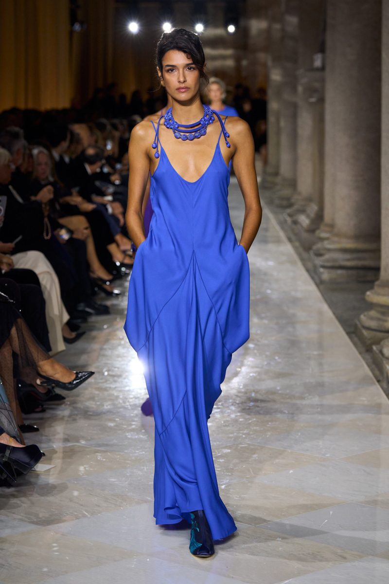 Model wearing a flowing cobalt blue silk gown with thin straps and a deep neckline, styled with a chunky blue statement necklace at Giorgio Armani runway show