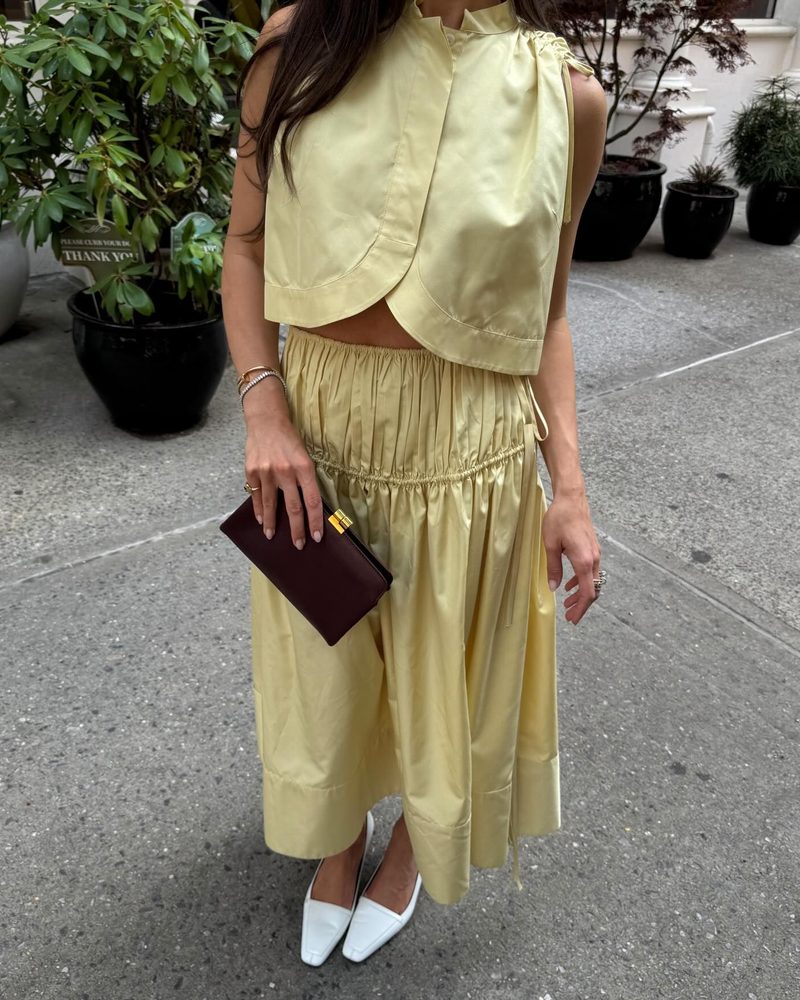 Butter yellow two-piece outfit featuring a cropped top and a flowy midi skirt%2C paired with white pointed-toe flats and a burgundy clutch