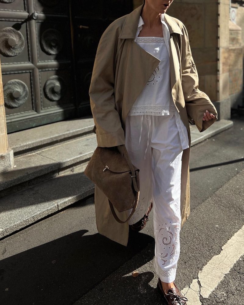 Light beige trench coat layered over a white embroidered linen top and matching pants%2C complemented by a taupe bag and brown lace-up shoes
