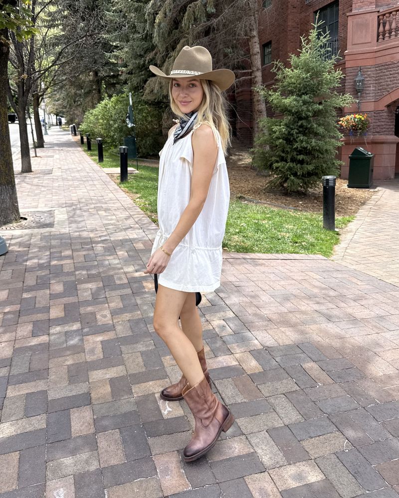 Woman wearing a sleeveless white summer dress paired with brown leather mid-calf boots%2C a tan wide-brimmed cowboy hat%2C and a black and white neck scarf on a city sidewalk