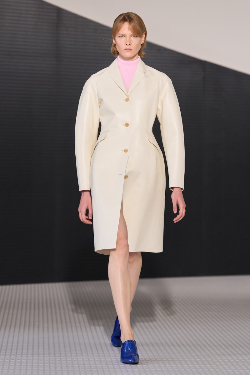 Model wearing Jil Sander cream leather tailored coat with structured shoulders, pink turtleneck, and blue loafers on runway