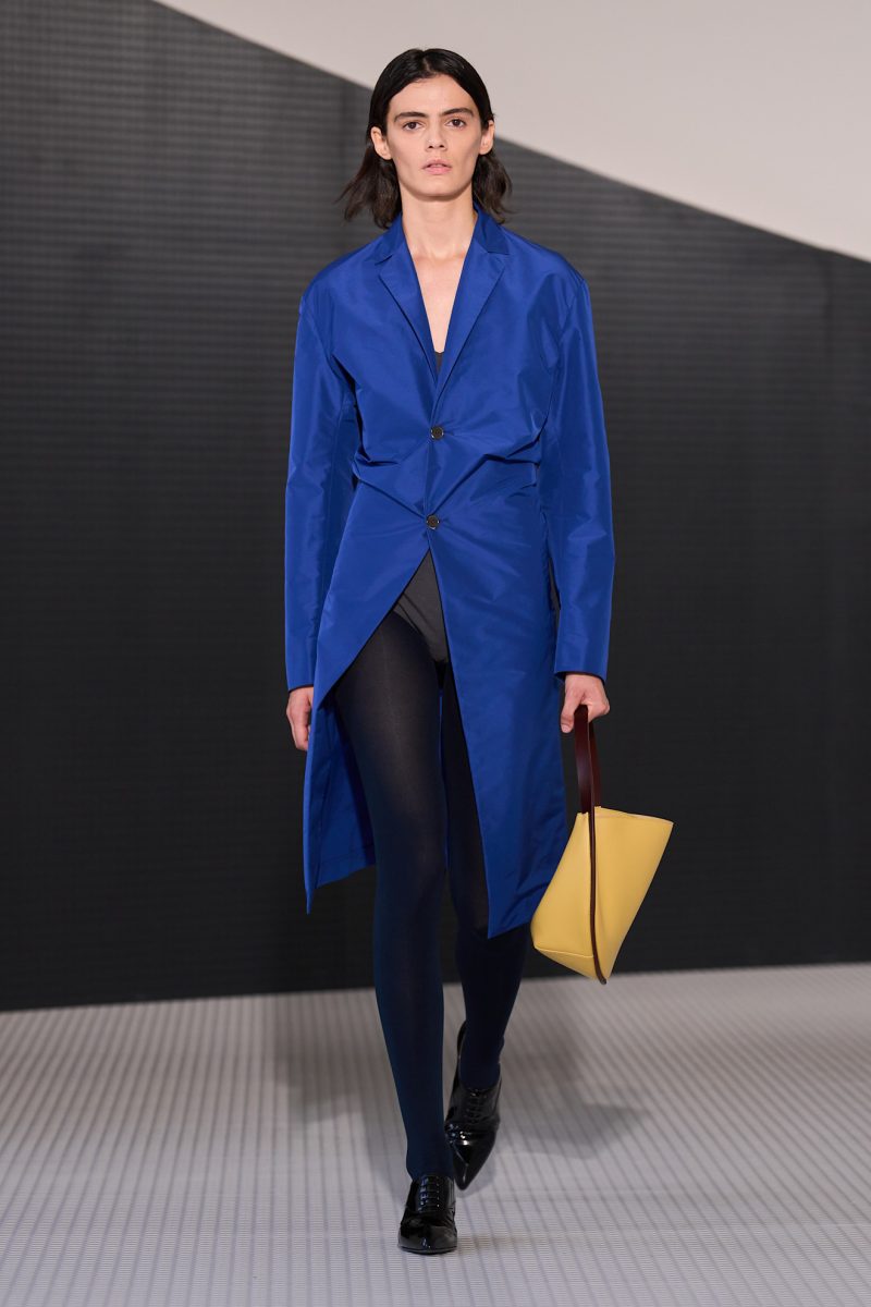 Model wearing a tailored blue Jil Sander coat with black leggings, black oxford shoes, and carrying a yellow handbag on runway