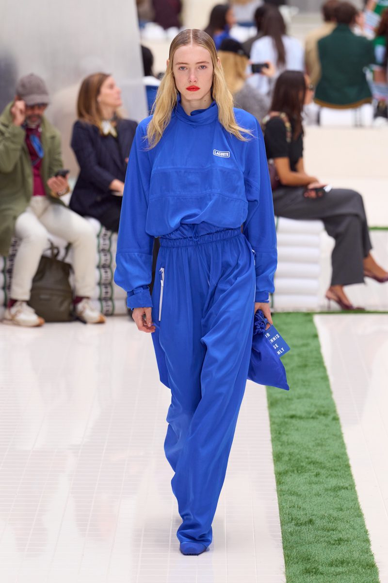 Model wearing a vibrant blue Lacoste tracksuit with a high-neck pullover and elastic waist pants, styled with a matching clutch bag