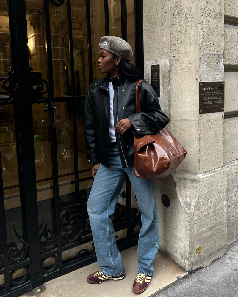 Woman wearing a black leather jacket%2C light blue jeans%2C beige beret%2C and brown leather tote bag standing by ornate black gate in urban setting