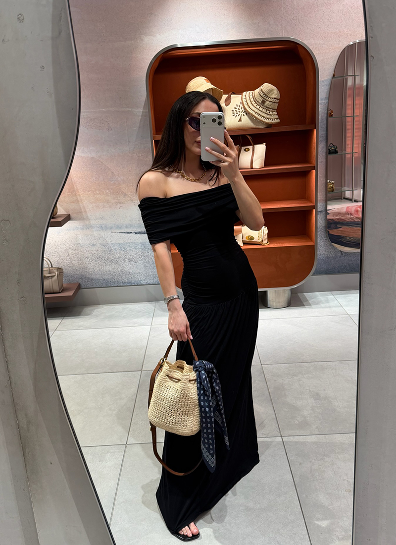 Black off-shoulder maxi dress with woven straw bucket bag, scarf detail and sandals styled in boutique fitting room mirror