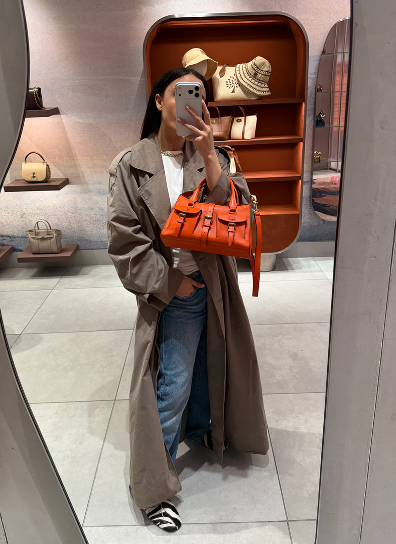 Beige trench coat over white tee and jeans with bright orange leather satchel, styled in boutique fitting room mirror