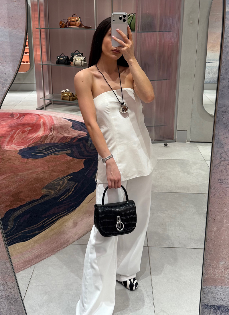 Strapless white top with wide-leg trousers, black top-handle bag and pendant necklace styled in boutique fitting room mirror
