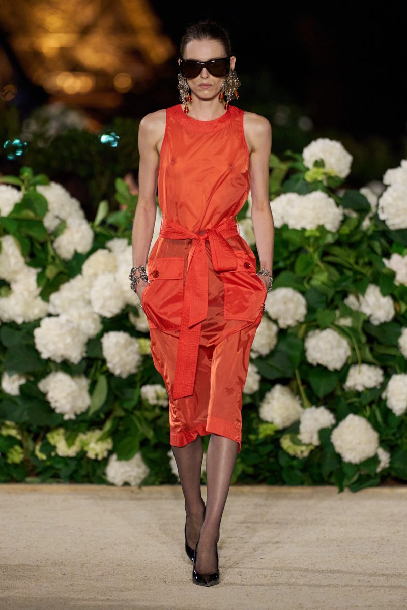 Saint Laurent sleeveless orange satin midi dress with oversized pockets and waist tie, styled with black sheer tights and statement earrings