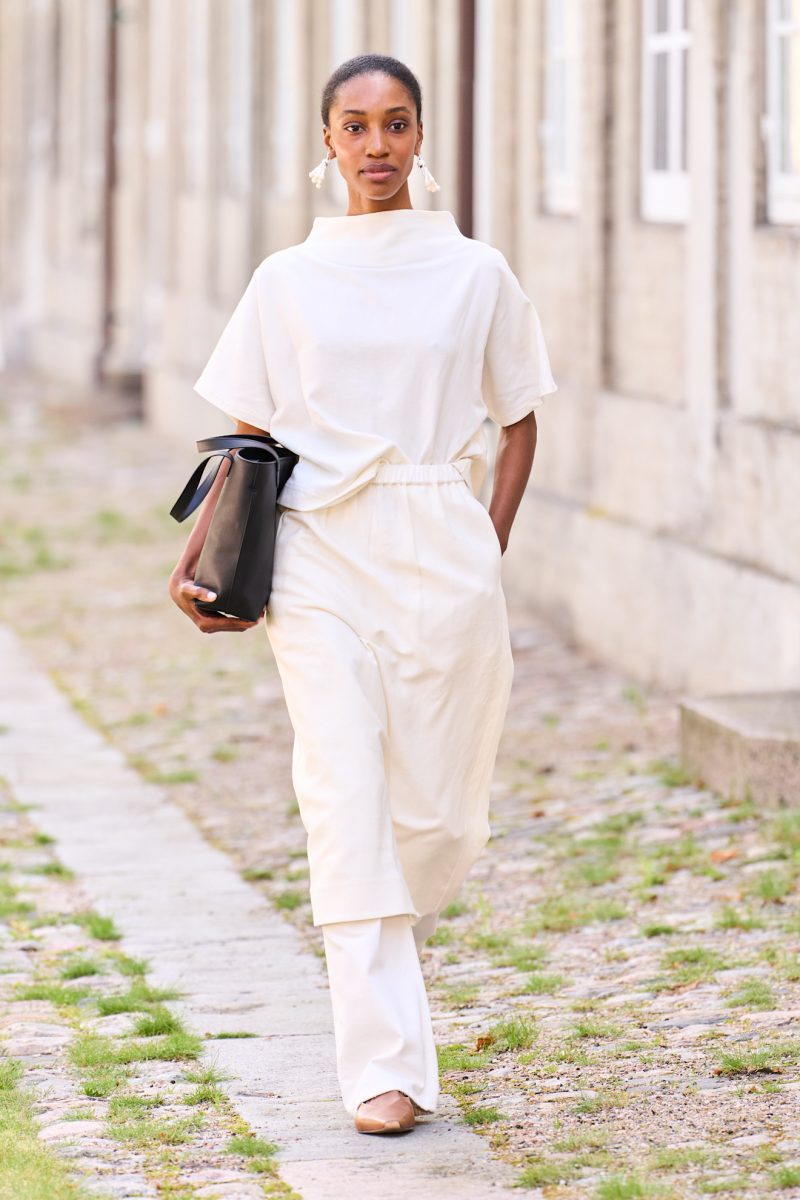 Model wearing Skall Studio cream oversized top and matching wide-leg pants with beige flats and black handbag on cobblestone street