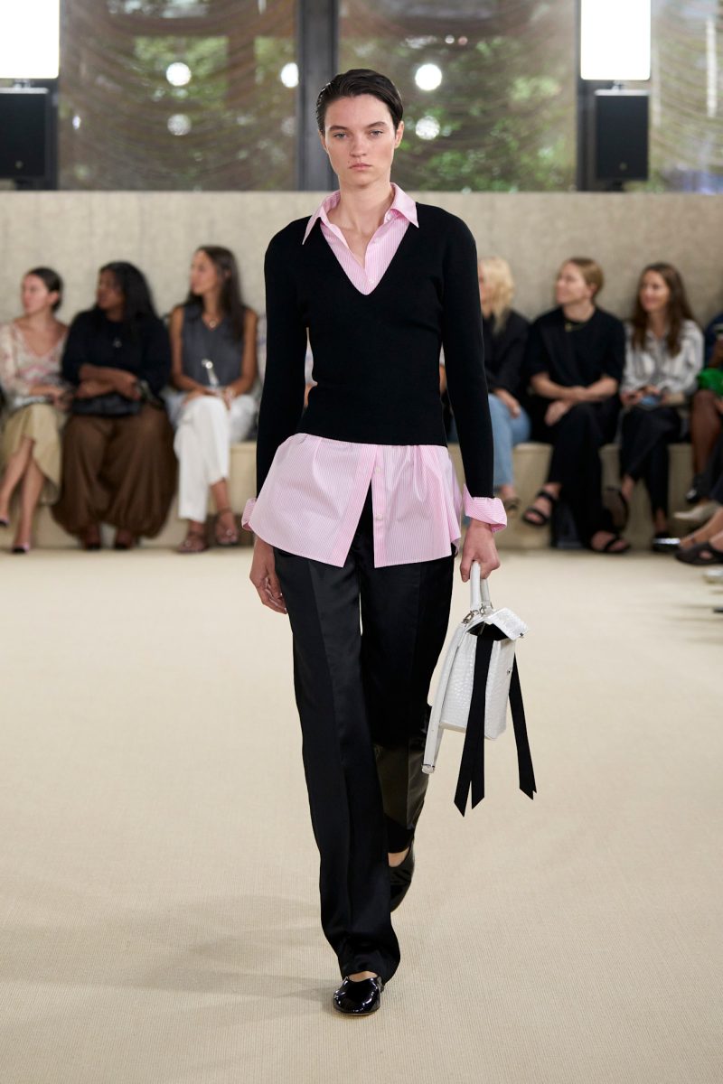 Model wearing Toteme spring 2026 black v-neck sweater layered over pink striped shirt with black tailored pants and white handbag with black ribbons
