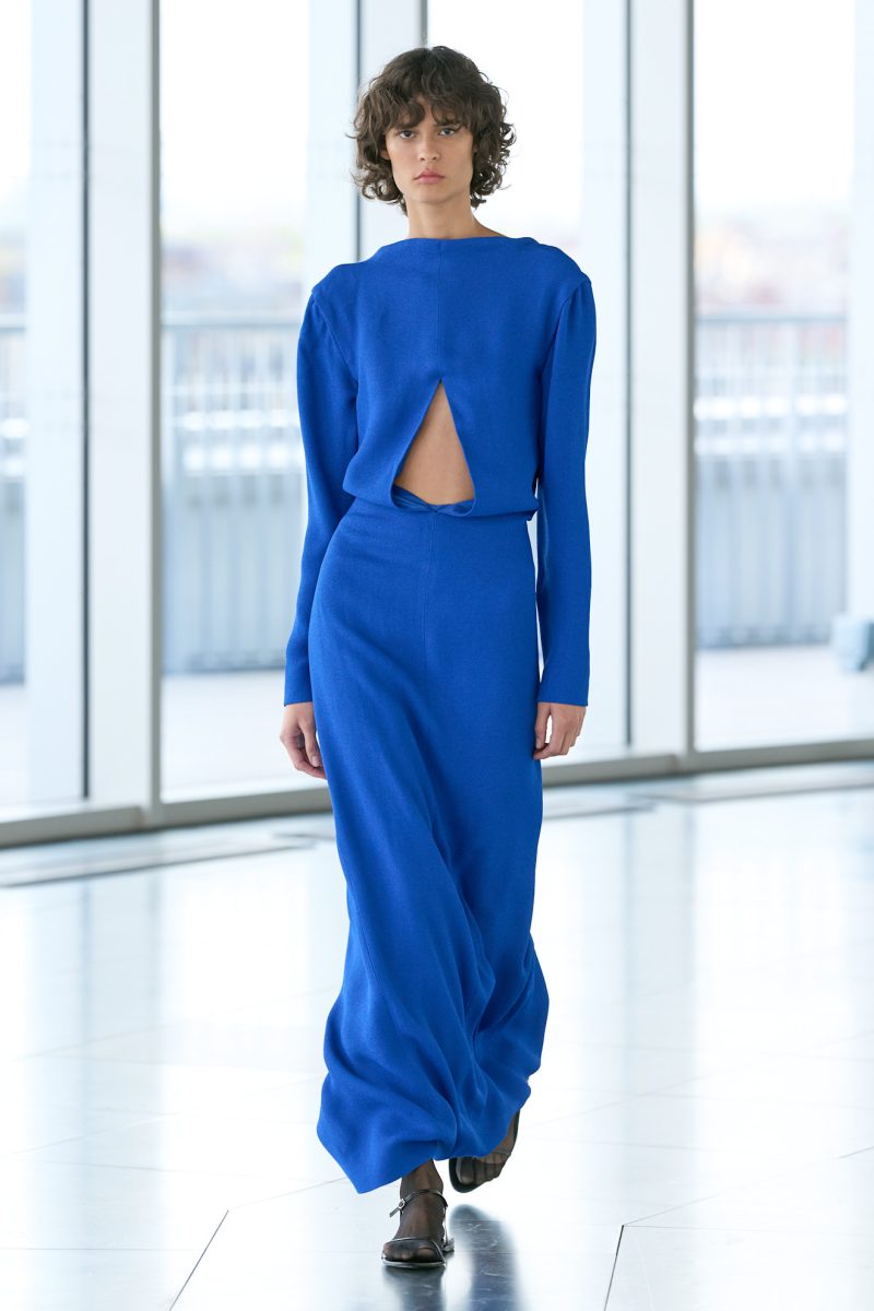Model wearing a vibrant blue long-sleeve knit dress with a back cutout detail and a flowing maxi skirt, styled with black strappy heels