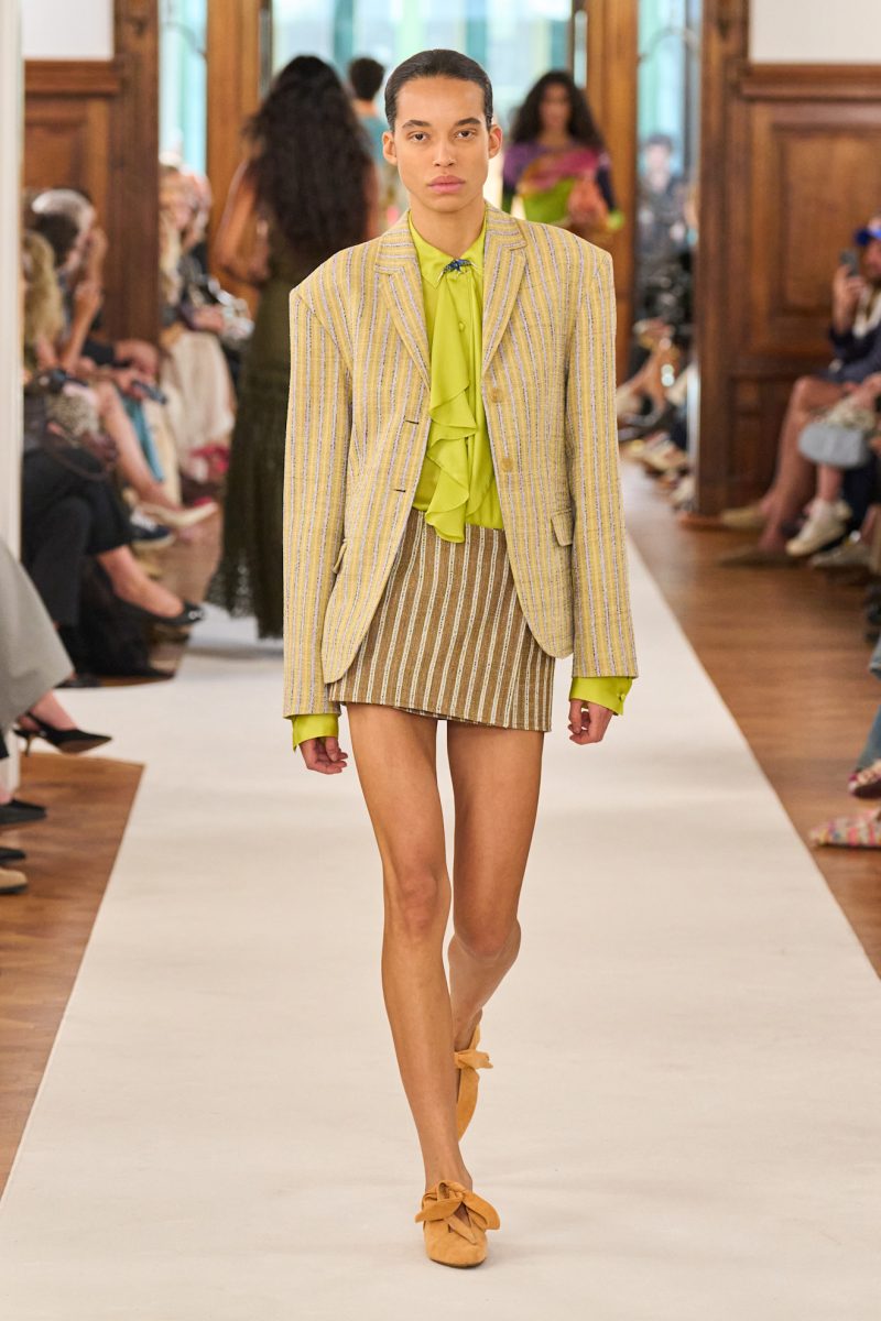 Model wearing Ulla Johnson spring 2026 striped beige blazer, lime green ruffled blouse, brown striped mini skirt, and tan suede bow flats on runway