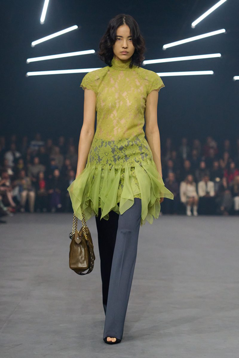 Model wearing Valentino spring 2026 green lace top with ruffled hem paired with two-tone tailored pants and brown handbag on runway