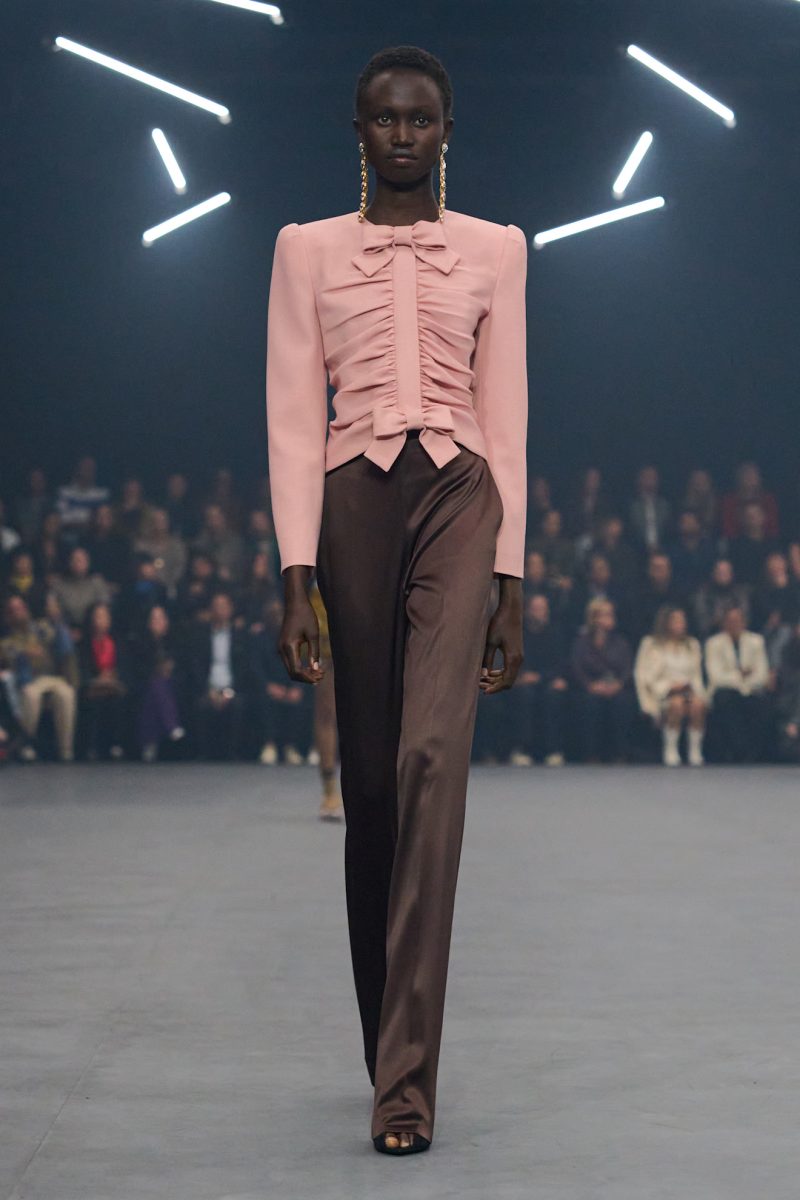 Model wearing Valentino spring 2026 pink ruched bow blouse paired with high-waisted brown satin trousers on runway